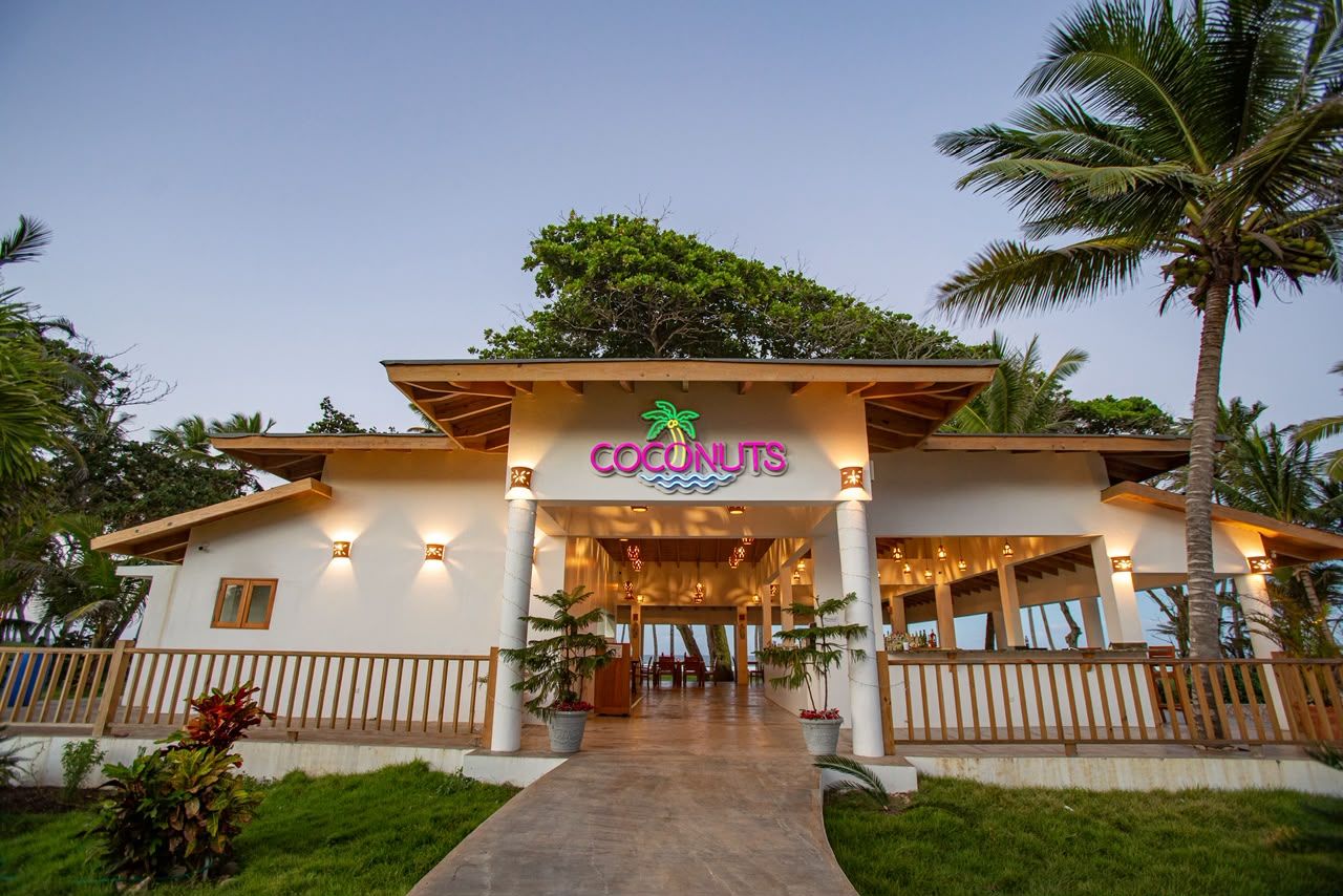Entrada de hotel com placa 'Coconuts', galeria coberta e palmeiras.