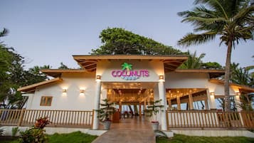 Entrada de hotel com placa 'Coconuts', galeria coberta e palmeiras.
