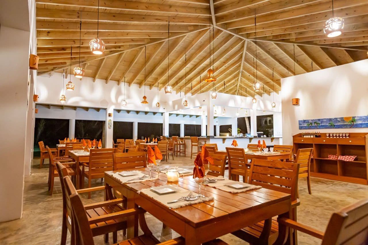 Uma ampla área de jantar com mesas e cadeiras de madeira, iluminação pendente e teto alto.