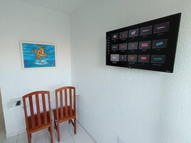 Quarto com duas cadeiras de madeira, televisão instalada na parede exibindo vários canais e um quadro na parede.