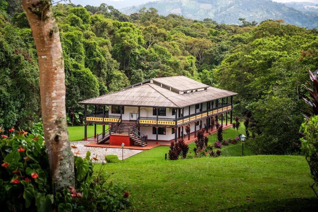 Um edifício de dois andares com varanda, cercado por vegetação exuberante e um tapete vermelho que leva à entrada.