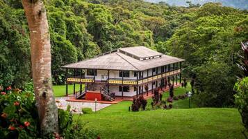 Um edifício de dois andares com varanda, cercado por vegetação exuberante e um tapete vermelho que leva à entrada.