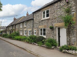 Uma fileira de casas de pedra com portas e janelas brancas, um caminho pavimentado e vegetação ao longo da calçada.