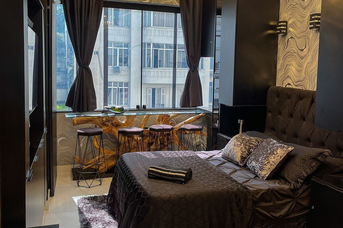 Quarto moderno com uma cama grande, um candelabro, uma escrivaninha com banquinhos e vista para prédios por grandes janelas.