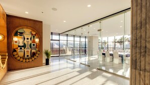 Um hall de hotel moderno com uma ampla parede de vidro, um balcão de recepção de madeira e vista para a paisagem urbana.