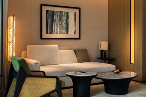 Quarto de hotel moderno com sofá, duas mesas laterais, uma cadeira, um abajur e uma obra de arte emoldurada na parede.