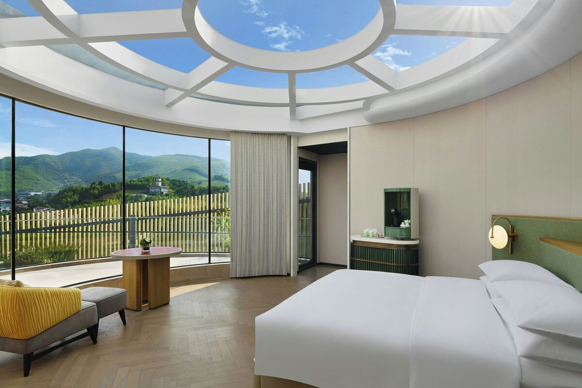 Quarto de hotel moderno com uma cama grande, uma mesinha, área de estar e vista para as montanhas por grandes janelas.
