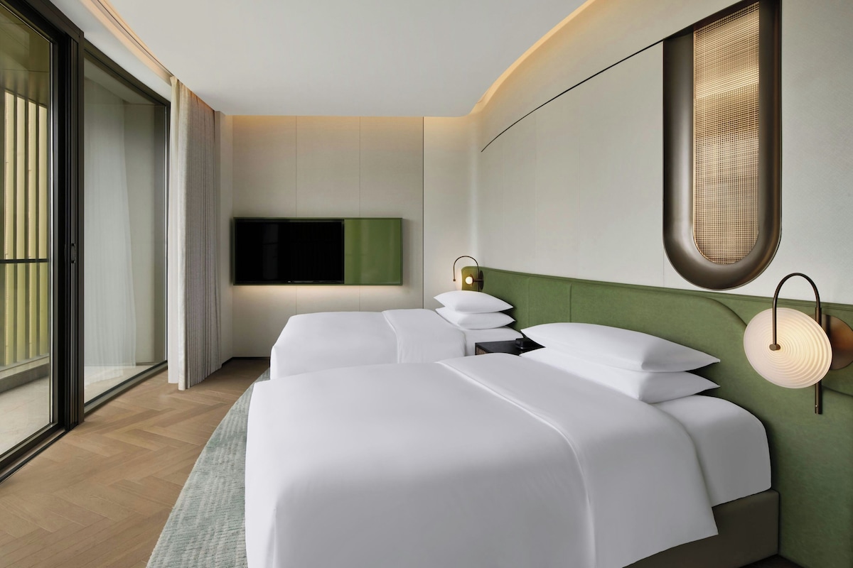 Quarto de hotel moderno com uma cama grande, televisão de tela plana na parede e uma janela ampla com cortinas.