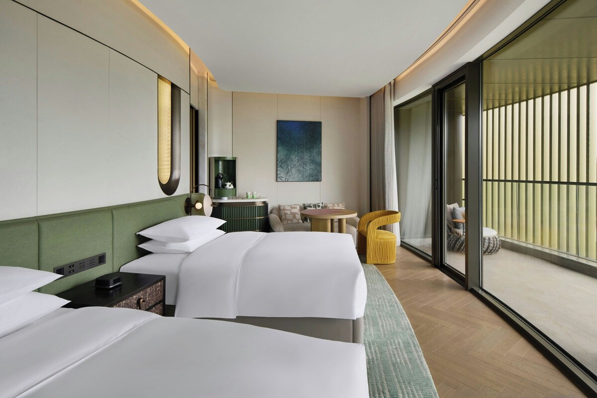 Quarto de hotel com duas camas, uma mesinha, uma cadeira, um sofá, um quadro e uma janela grande.