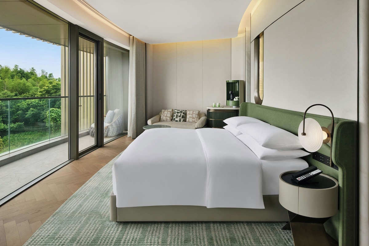 Quarto de hotel moderno com uma cama grande, uma mesinha de centro, um criado-mudo e vista para o verde através da varanda.