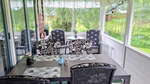 Uma área de varanda coberta com mesa e cadeiras, janelas amplas e vista para o verde.