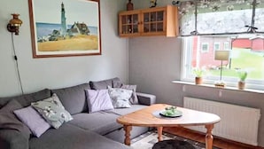 Uma sala de estar com um sofá cinza, uma mesa de centro de madeira, um banquinho redondo, um quadro emoldurado, um armário e uma janela com plantas.