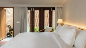 Quarto de hotel com uma cama grande, uma cadeira de rodas e vista para uma parede de tijolos e vegetação.