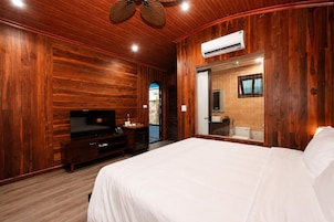 Quarto com paredes de madeira, uma cama, uma televisão, banheiro com espelho e aparelho de ar condicionado.