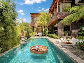 Uma piscina com um espaço de refeições flutuante, cercada por vegetação exuberante e um edifício moderno com grandes janelas.