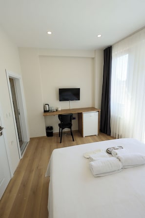 Quarto de hotel com cama, escrivaninha com computador, cadeira, mini-frigorífico e televisão na parede.