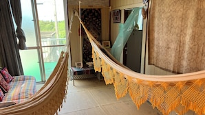 Quarto com rede, cama com roupa de cama estampada, mesa pequena com porta-retrato e vista para o verde lá fora.