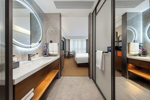 Quarto de hotel com um espelho grande, uma penteadeira de madeira, uma cama e vista para o saguão do hotel.