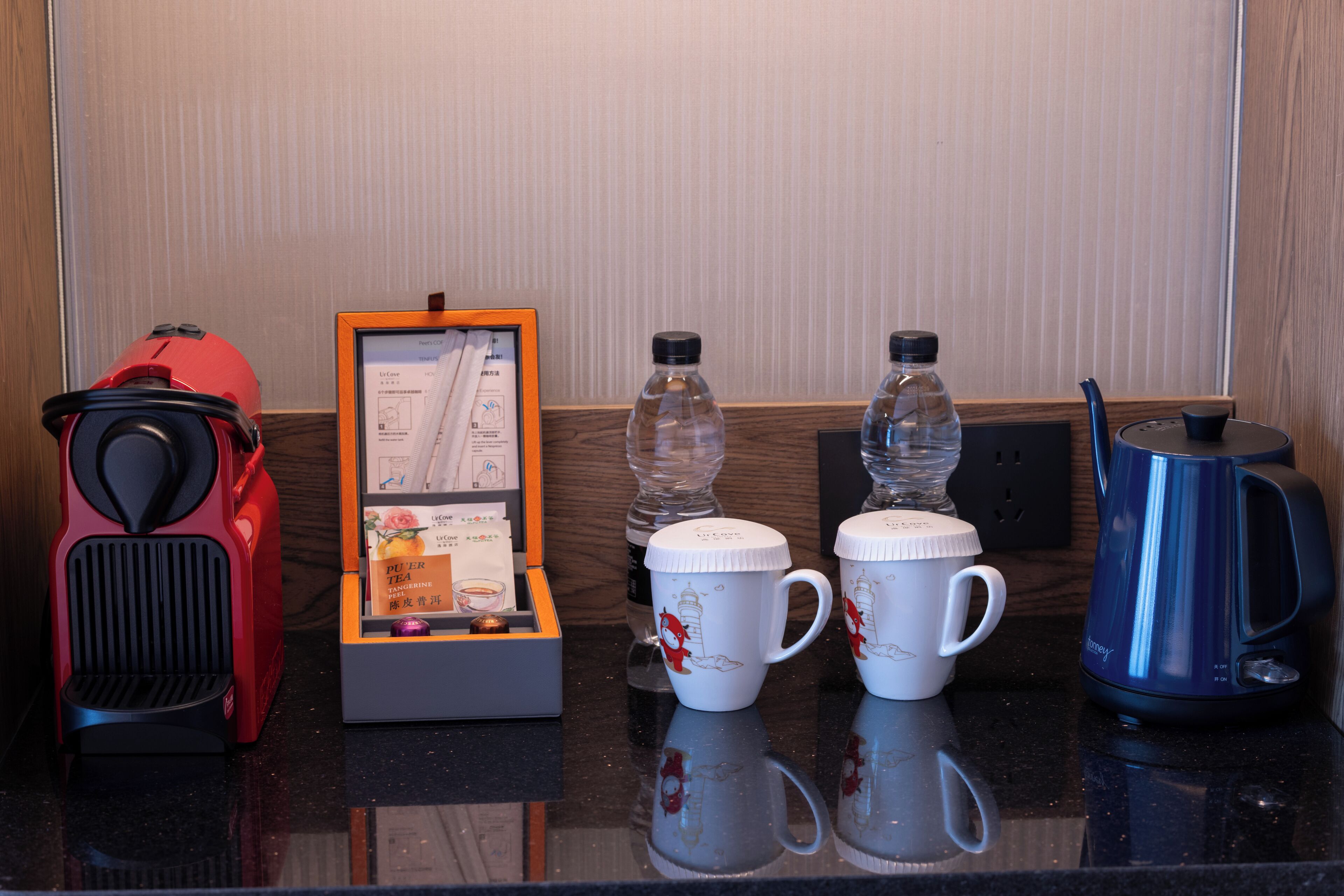 Balcão de quarto de hotel com cafeteira, uma caixa de chá, duas canecas e uma garrafa de água.