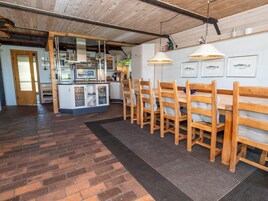 Interior de restaurante com cadeiras de madeira, uma mesa de jantar central, área de cozinha e chão de tijolos.