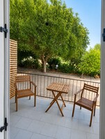 Um pátio externo com duas cadeiras de madeira e uma mesa, um cercado de madeira treliçada e uma árvore ao fundo.