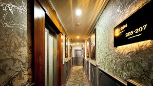 Um corredor com paredes de mármore e portas de madeira, que leva a um quarto com um espelho grande e uma placa com números.