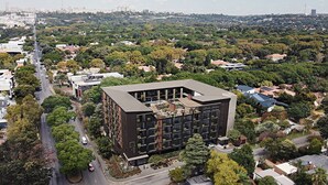 Um edifício com vários andares e telhado plano, rodeado por vegetação e áreas residenciais.