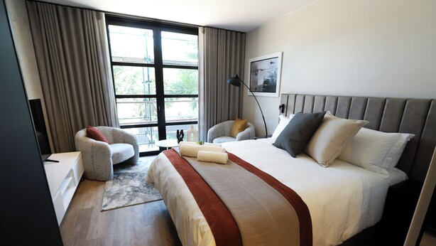 Quarto de hotel com cama, duas poltronas, escrivaninha, abajur e janela com cortinas.