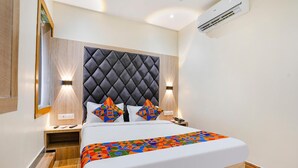 Quarto de hotel com cabeceira estofada, cama com colcha de padrão geométrico colorido, mesas de cabeceira com luminárias e um aparelho de ar condicionado.
