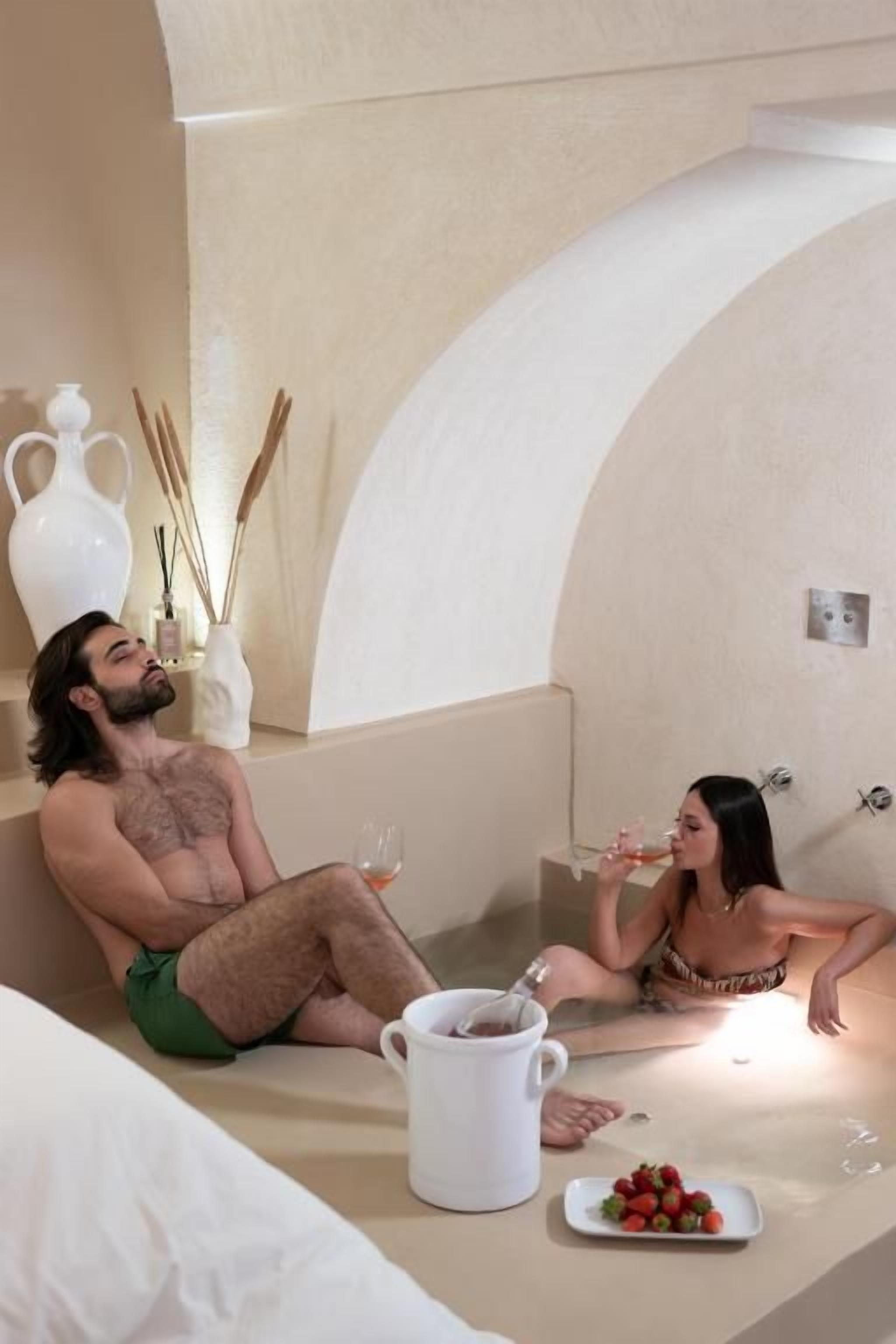Um casal relaxando em uma banheira com um jarro branco e morangos em um prato.