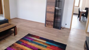 Quarto com piso de madeira, tapete listrado e colorido, sofá, porta com painel de vidro e área de refeições visível através de uma porta aberta.