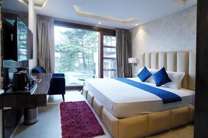 Quarto de hotel com cama, escrivaninha com cafeteira, cadeira, TV e vista para árvores pela janela.
