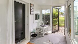 Quarto com porta de vidro deslizante para um jardim, uma cadeira branca, um escorregador de metal para sapatos e uma pequena mesa com uma garrafa.