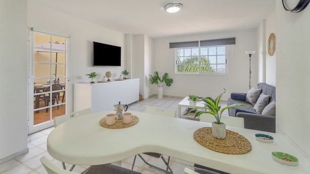 Uma sala de estar moderna com televisão de tela plana, um sofá, uma mesa de jantar com cadeiras e uma área de cozinha com geladeira e plantas em vasos.