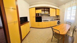 Uma cozinha compacta com armários amarelos, micro-ondas, forno e uma área de refeições com mesa de madeira e cadeiras de metal.