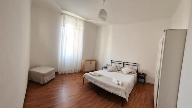 Um quarto com piso de madeira, uma cama com roupa de cama branca, uma mesinha de cabeceira e uma janela com cortinas.