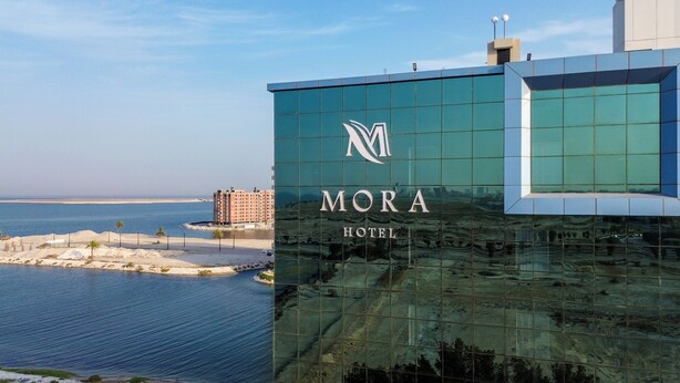 Um hotel moderno com fachada de vidro refletivo, logotipo em destaque e a inscrição 'MORA HOTEL'.