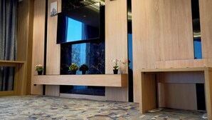 Um lobby moderno com uma televisão grande, painéis de madeira, uma parede de destaque em mármore e plantas em vasos.