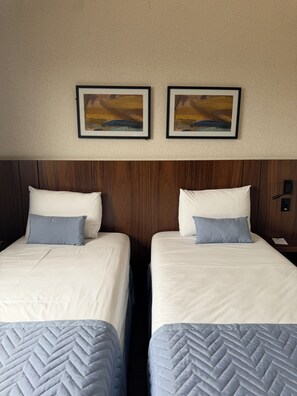 Quarto de hotel com duas camas, cada uma com travesseiro e edredom acolchoado. Há duas pinturas emolduradas na parede acima das camas.