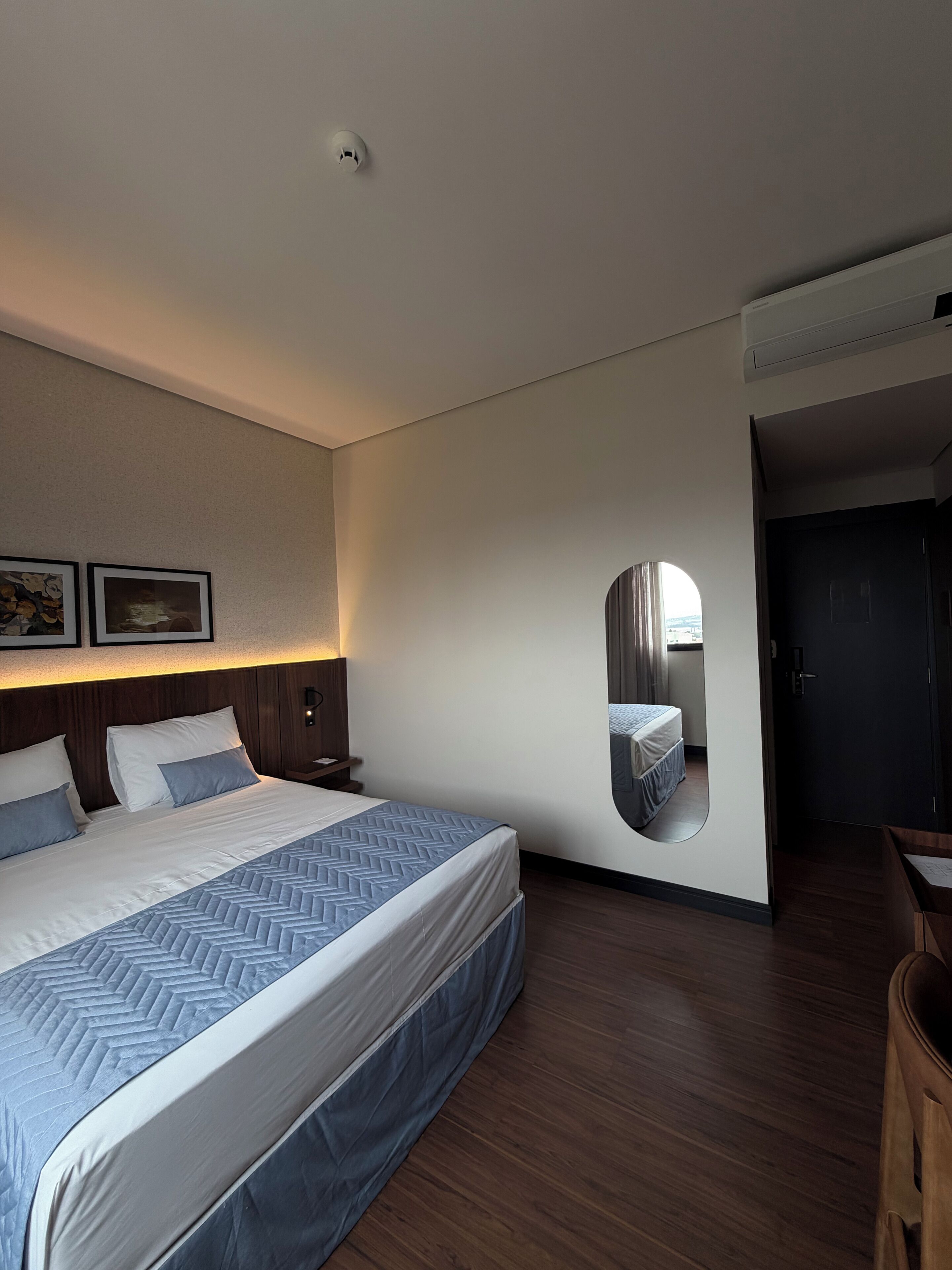 Quarto de hotel com uma cama, um espelho, chão de madeira e uma porta.
