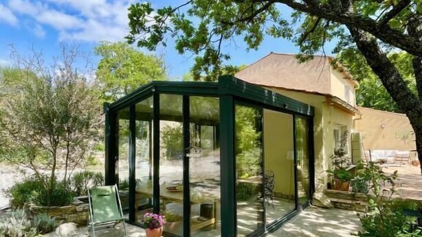Estrutura envidraçada com teto, rodeada por um jardim com plantas em vasos e uma cadeira.