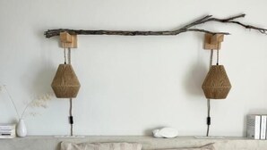 Uma cama com colcha bege estampada, duas almofadas e luminária fixada na parede com cúpula de vime e detalhes em galho.