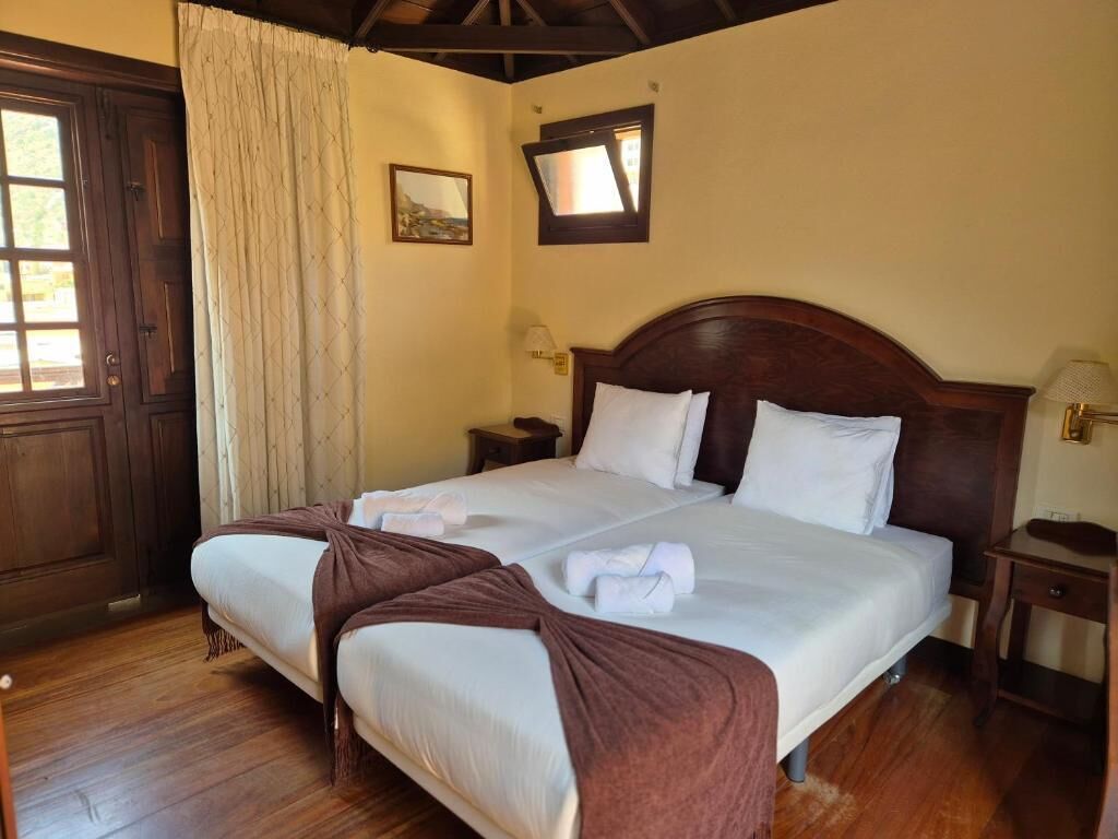 Uma cama de casal com lençóis brancos, um cobertor marrom, duas mesas de cabeceira com luminárias, uma porta de madeira e uma janela com cortinas.