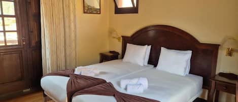 Uma cama de casal com lençóis brancos, um cobertor marrom, duas mesas de cabeceira com luminárias, uma porta de madeira e uma janela com cortinas.