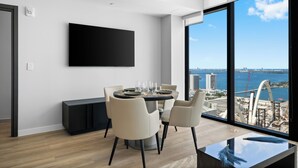 Uma sala de jantar moderna com uma mesa redonda, cadeiras, uma televisão de tela plana e uma grande janela com vista para a paisagem urbana e uma ponte.