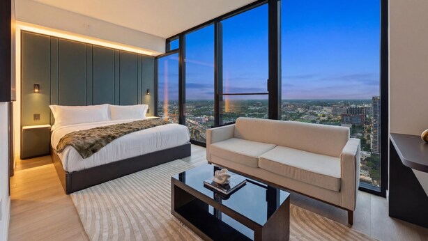 Quarto moderno com cama grande, sofá, mesa de centro e vista da cidade através de amplas janelas.