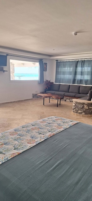 Uma sala de estar com sofá, mesa de centro, tapete estampado, parede de pedra e vista para o mar através de uma janela.