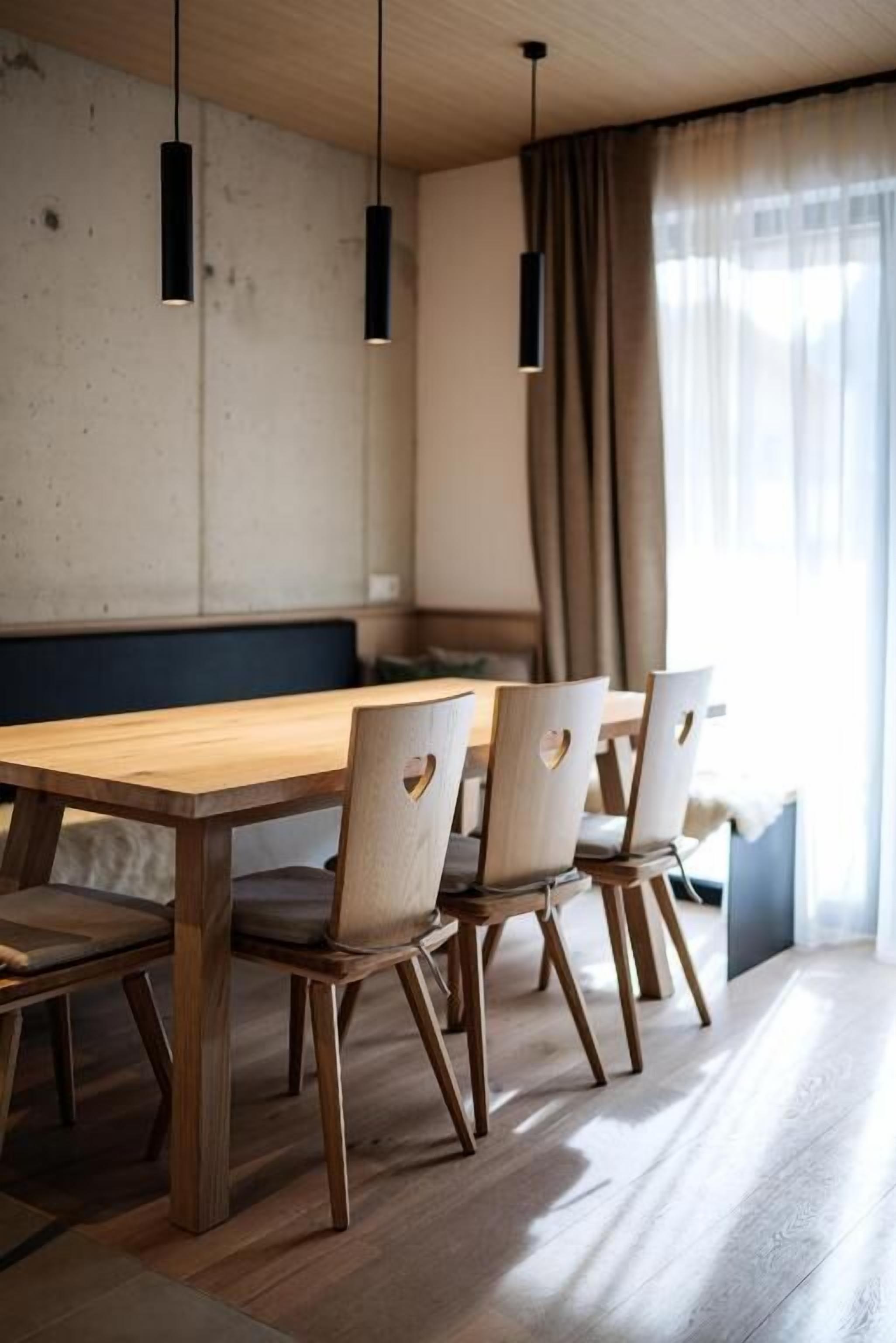 Uma área de refeições com mesa e cadeiras de madeira, luminárias pendentes e vista para o exterior através de uma janela com cortinas.