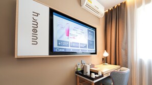 Quarto de hotel com televisão fixada na parede exibindo um anúncio de cuidados com a pele, uma escrivaninha com cadeira, uma garrafa de água e um abajur.