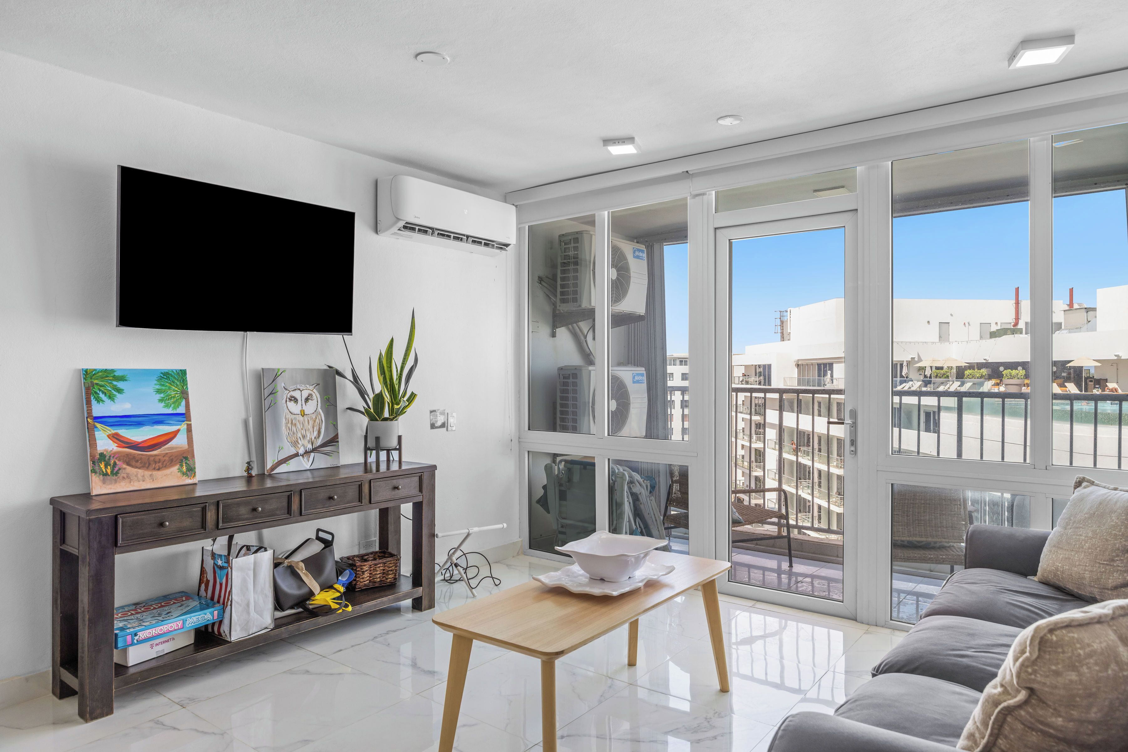 Uma sala de estar moderna com televisão de tela plana, mesa de console de madeira, sofá e janelas amplas com vista para a cidade.